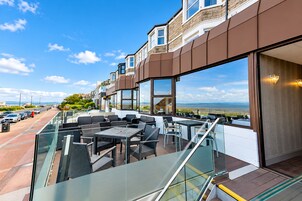 Terraço com mesas ao ar livre, vista para o mar e um edifício com grandes janelas.