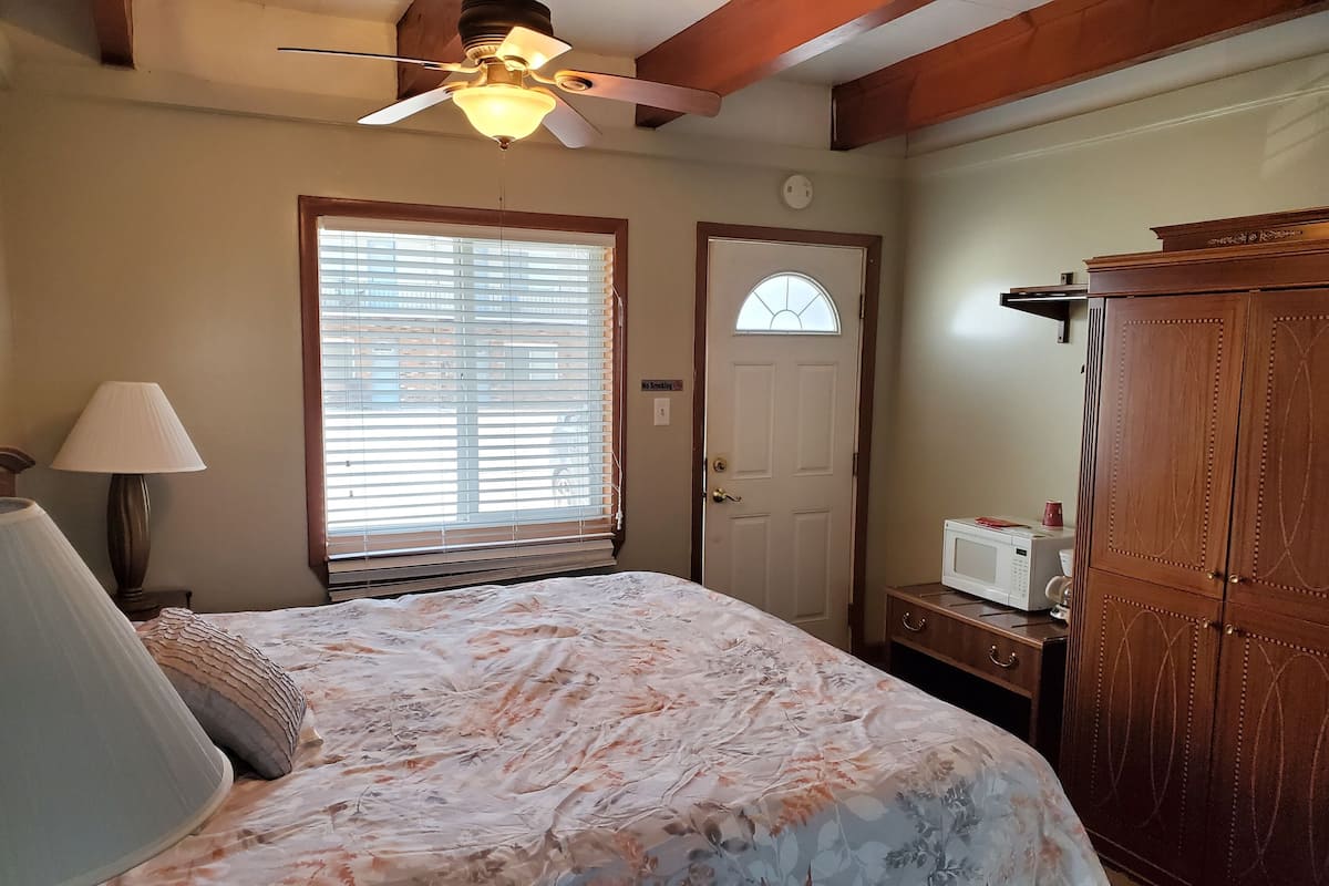 Quarto com cama, ventoinha no teto, janela com persianas, porta, guarda-roupa de madeira e mesa de cabeceira com abajur.