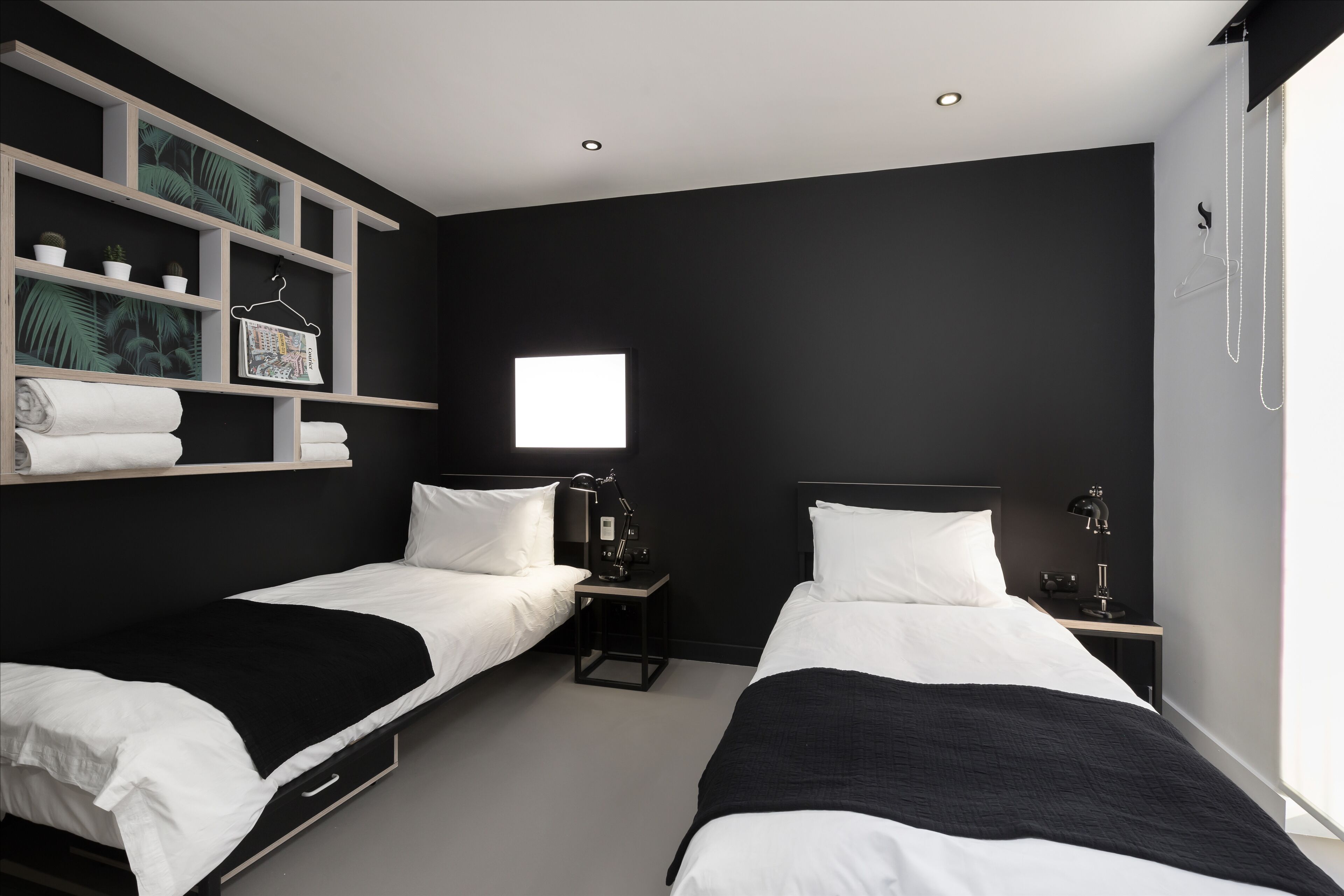 Quarto de hotel moderno com duas camas de solteiro, uma pequena mesa de trabalho, uma cadeira, prateleira com objetos decorativos e janela que proporciona luz natural.