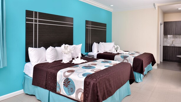 Quarto de hotel com duas camas, cada uma com cabeceira, um banheiro com pia e espelho, e uma pequena cozinha.