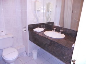Casa de banho com sanita branca, lavatório com duas torneiras, espelho e porta-toalhas.
