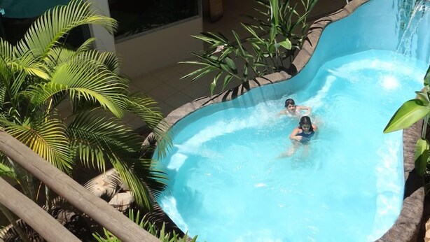 Piscina com duas pessoas na água, rodeada por plantas tropicais.