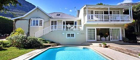 Uma casa de dois andares com piscina, varanda e montanhas ao fundo.