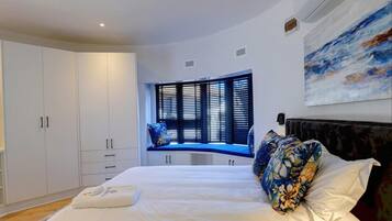 Quarto com uma cama grande, um banco embutido com assento, uma janela com persianas e um quadro na parede.
