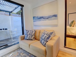 Uma sala de estar com um sofá bege, duas almofadas estampadas, um quadro na parede e vista através de uma porta de vidro.