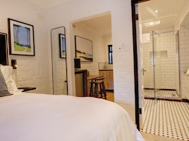 Quarto de hotel com uma cama grande, uma escrivaninha, uma cadeira e área de chuveiro com divisória de vidro.