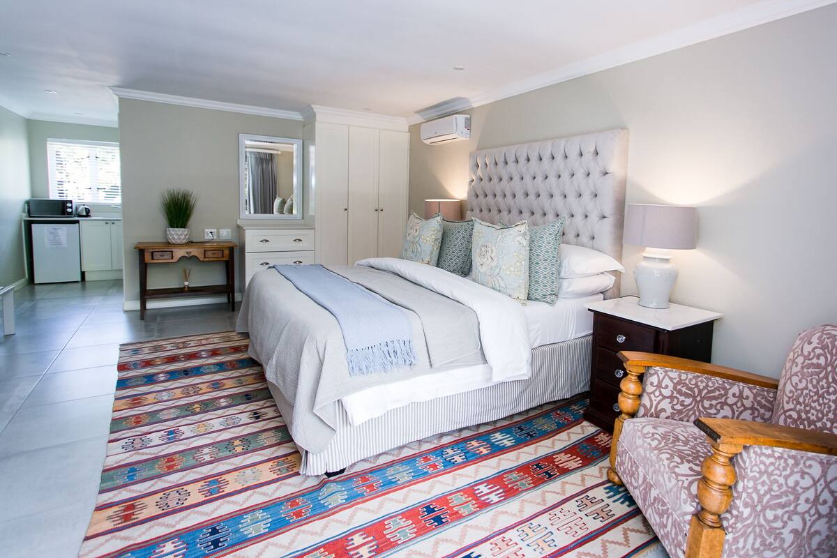 Quarto com uma cama grande, mesa de cabeceira com luminária, uma cadeira, cômoda e janela com vista para o exterior.