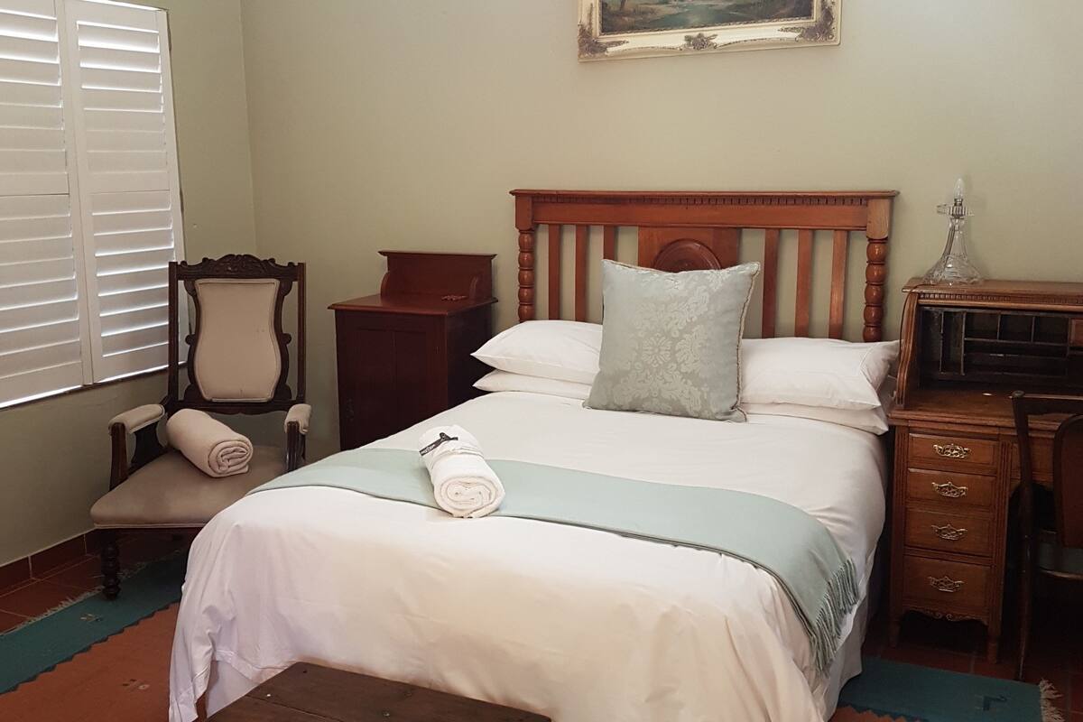 Uma cama bem arrumada com cabeceira de madeira, um pequeno baú de madeira, uma cadeira, uma mesinha de cabeceira e um quadro na parede.