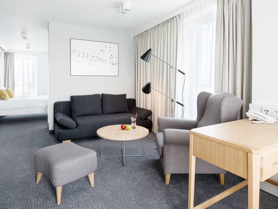 Quarto de hotel moderno com sofá cinza, mesa de madeira redonda, poltrona cinza e um pequeno banquinho.
