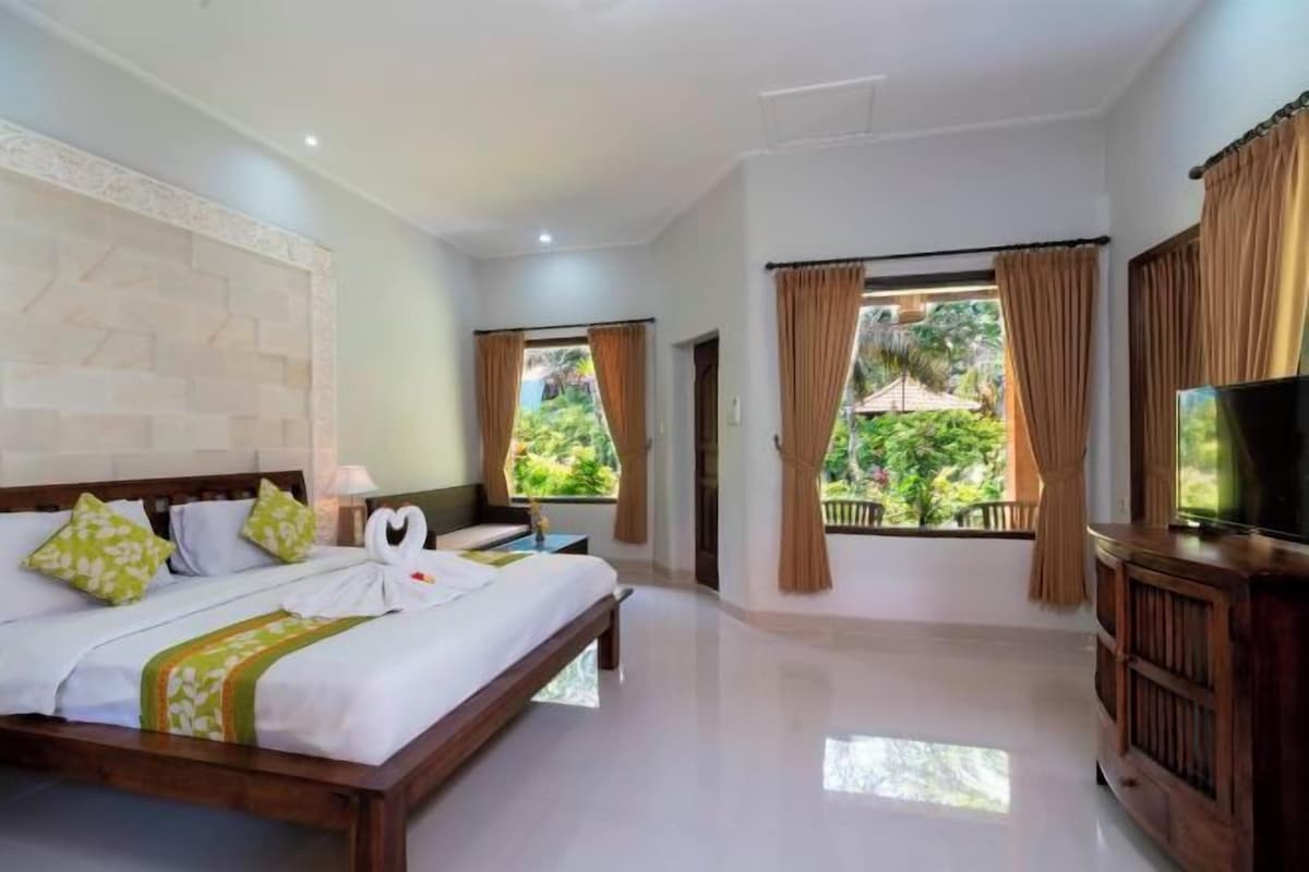 Quarto de hotel com cama, televisão, armário de madeira e vista para o verde pelas janelas.