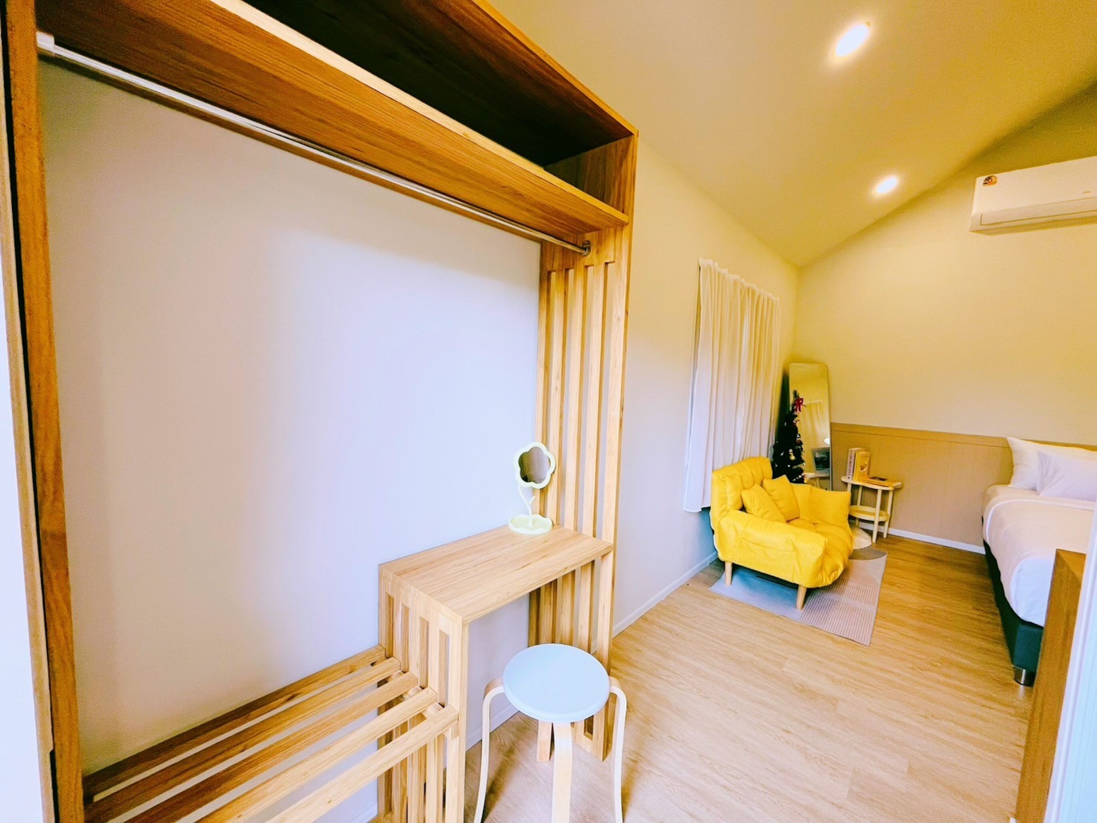 Um quarto com uma escrivaninha de madeira, um banquinho pequeno, um sofá amarelo, uma cama branca e um espelho.