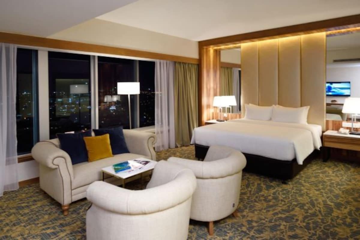 Quarto de hotel com uma cama grande, duas poltronas, uma mesa pequena e vista para a cidade à noite.