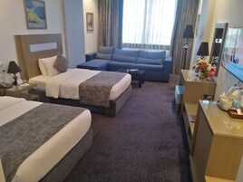 Quarto de hotel com duas camas, um sofá, uma mesa de trabalho com abajur e vista para o exterior.