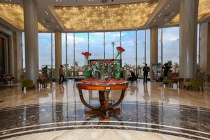 Um hall amplo com piso de mármore polido, uma mesa redonda central com flores vermelhas e grandes janelas com vista para palmeiras e um horizonte urbano distante.