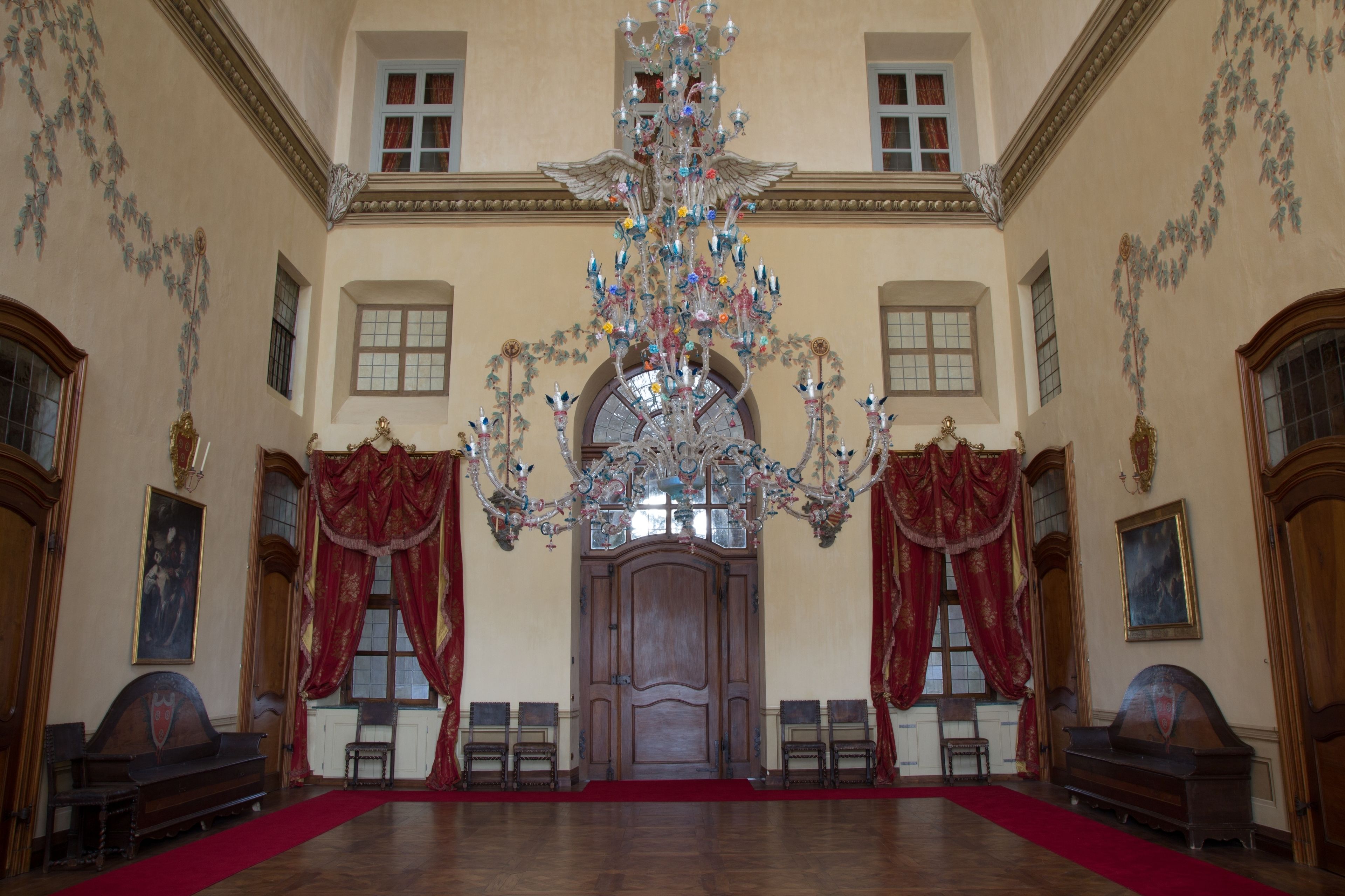 Um salão imponente com um grande candelabro, bancos de madeira e cortinas vermelhas.