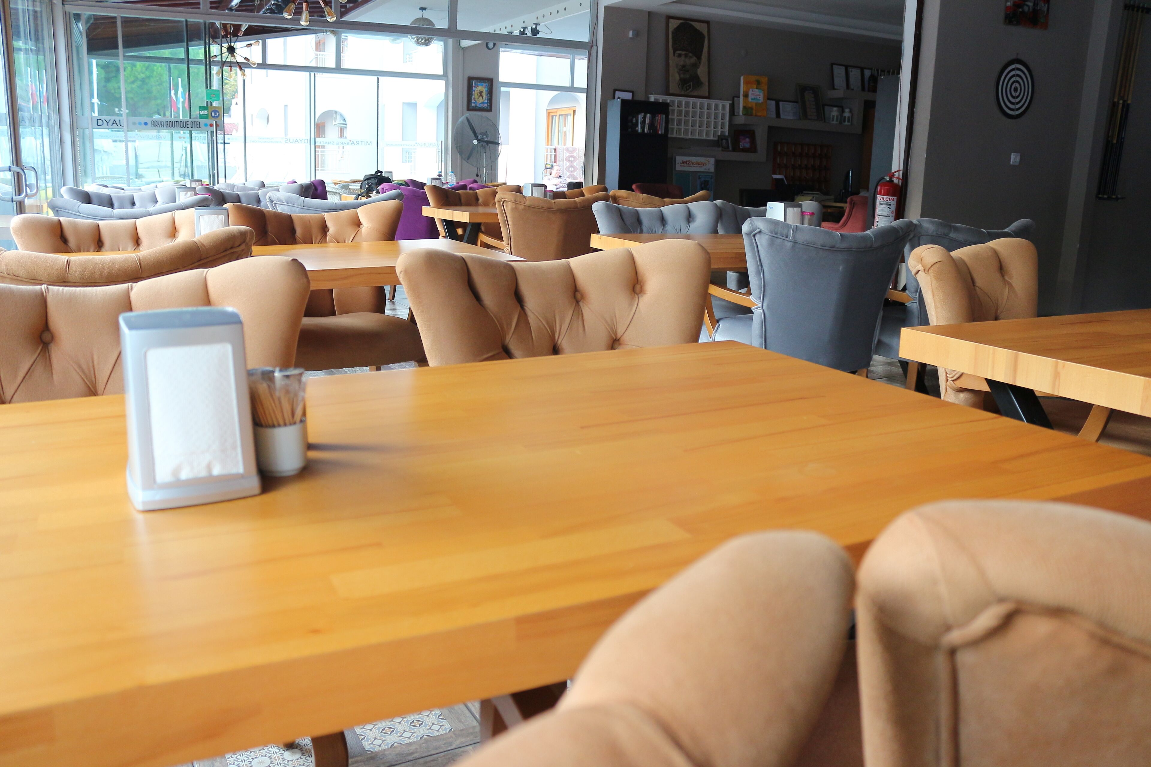 Um restaurante com mesas de madeira e cadeiras estofadas, grandes janelas e uma área externa visível.