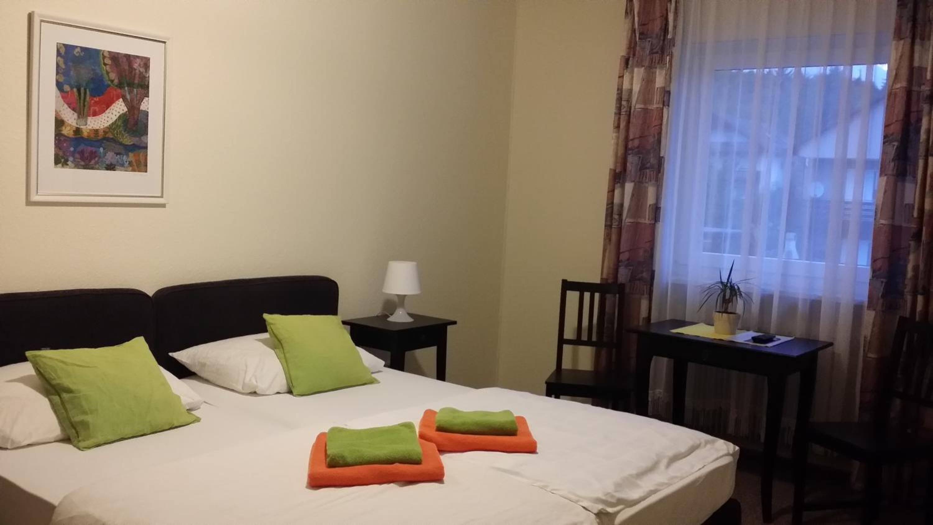 Quarto de hotel com cama, duas almofadas, mesa de cabeceira com abajur, cadeira, pequena mesa com uma planta e janela com cortinas.