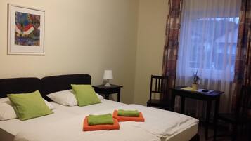Quarto de hotel com cama, duas almofadas, mesa de cabeceira com abajur, cadeira, pequena mesa com uma planta e janela com cortinas.