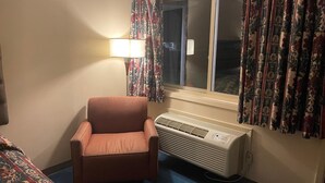 Quarto de hotel com uma cadeira, janela com cortinas, ar condicionado fixado na parede e uma luminária.