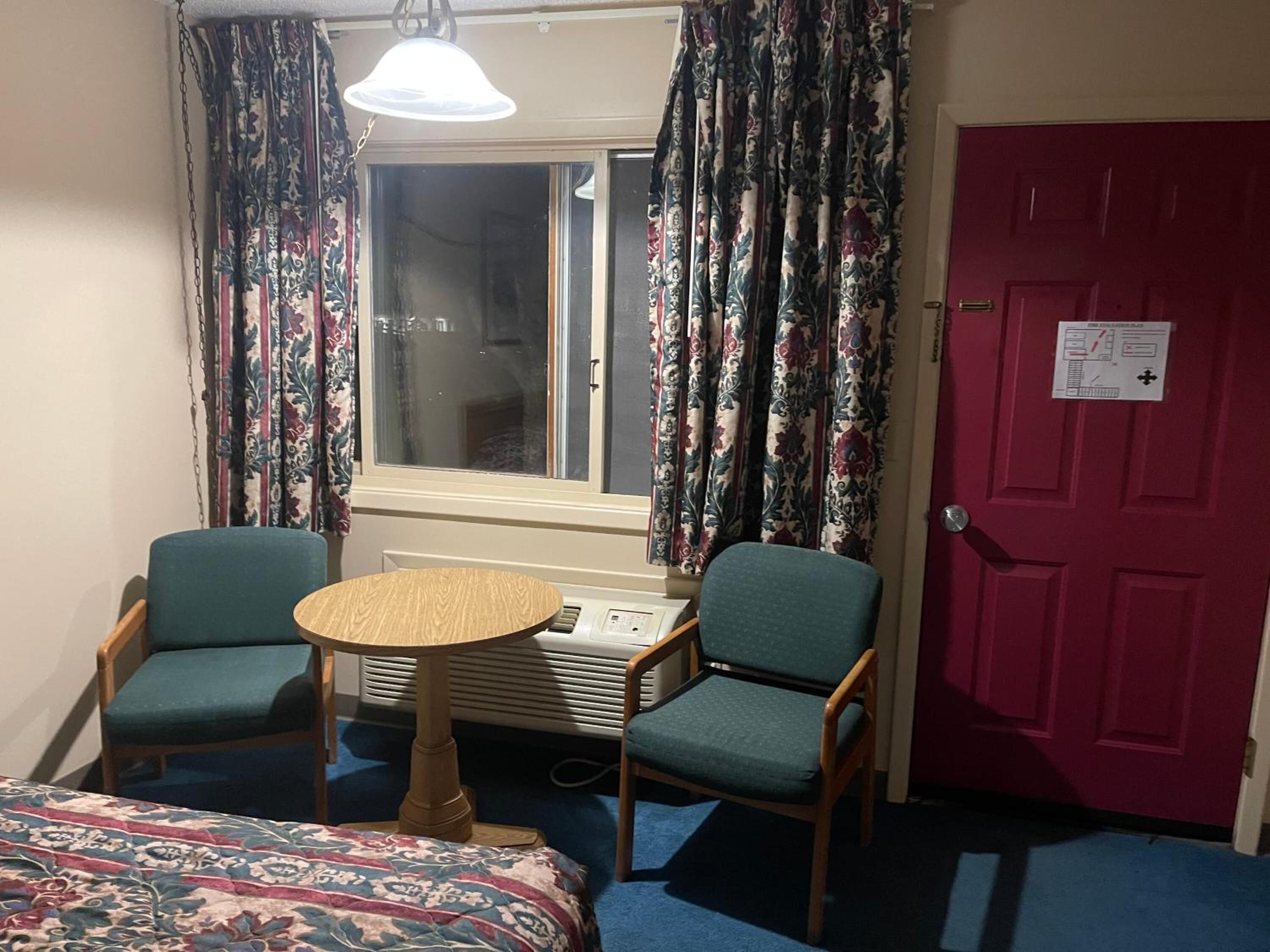 Quarto com duas cadeiras, uma mesa redonda pequena, cama com colcha estampada, janela com cortinas e porta vermelha.