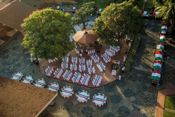 Uma área externa com mesas e cadeiras redondas, cercada por árvores e um gazebo central.
