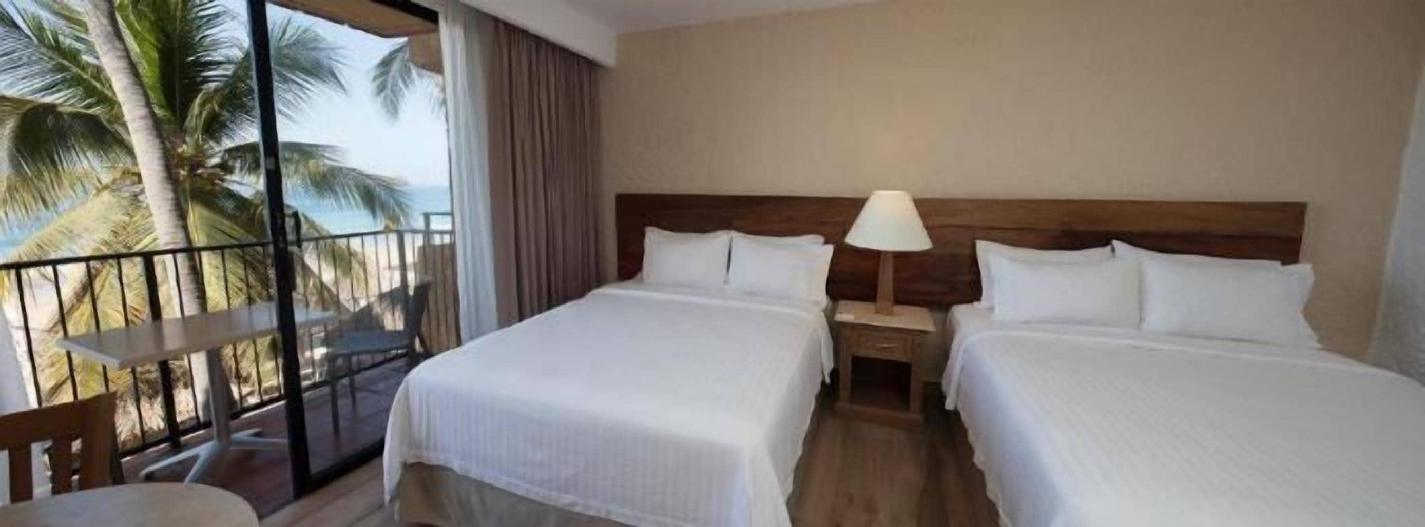 Quarto de hotel com duas camas, uma varanda e vista para palmeiras, e cabeceira de madeira.