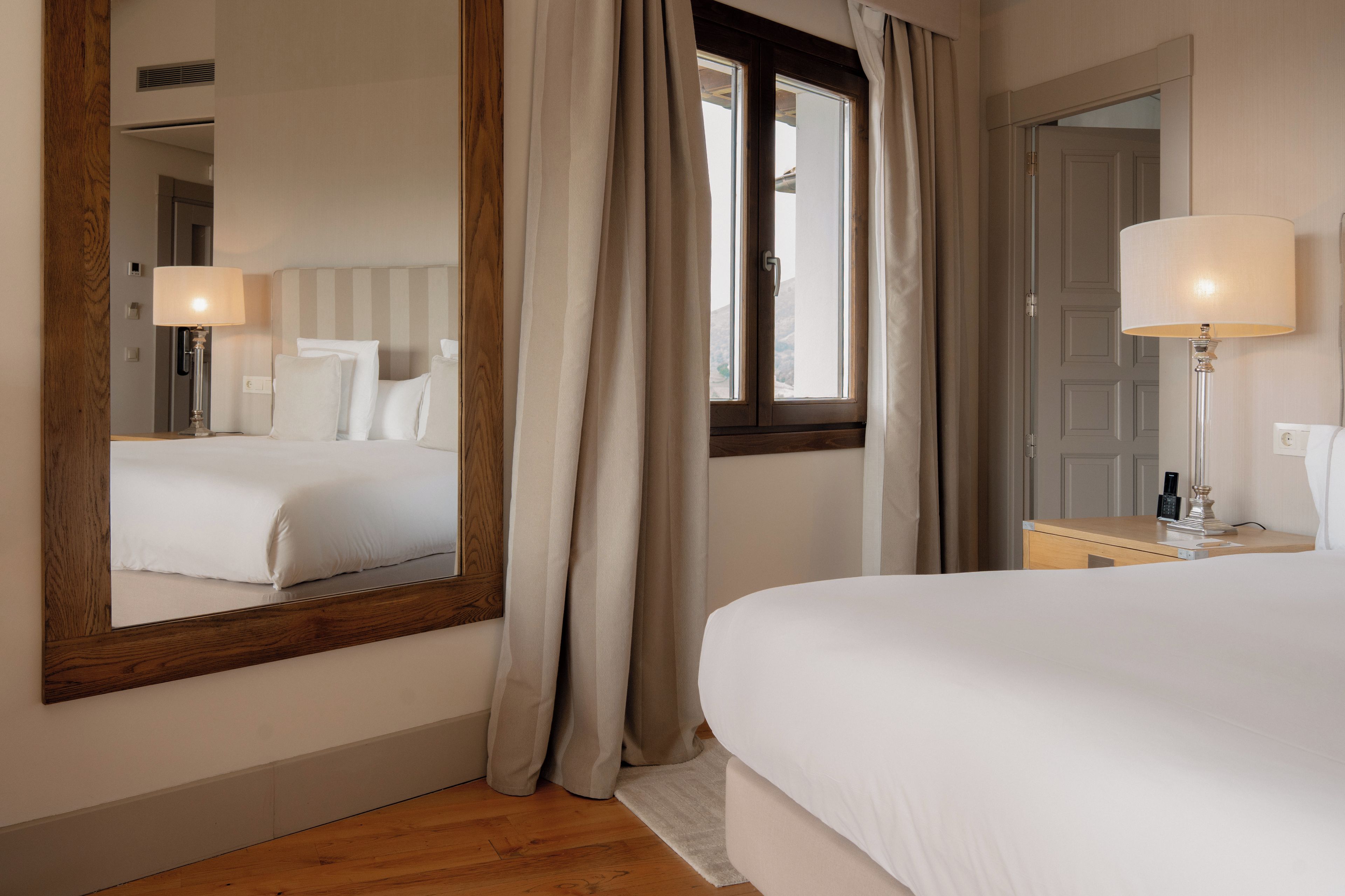 Quarto de hotel com uma cama grande, mesa de cabeceira com luminária, espelho e janela com cortinas.