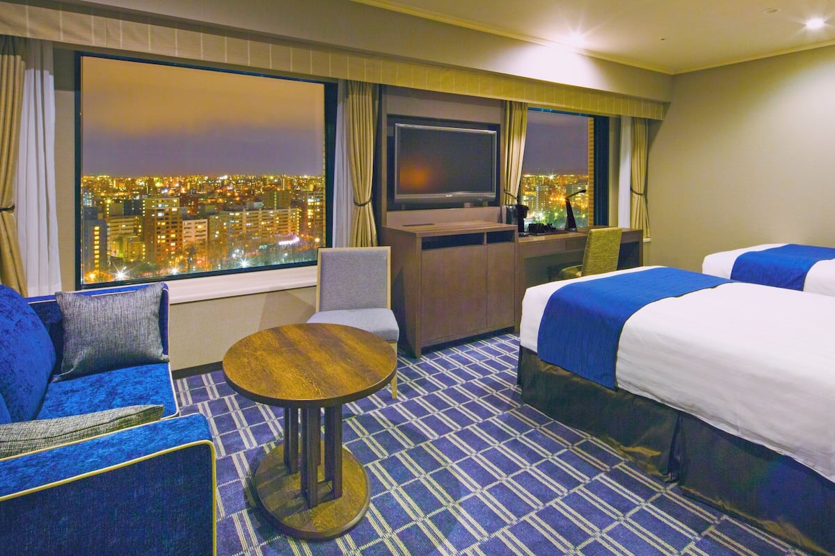 Quarto de hotel com duas camas, uma escrivaninha, uma cadeira, uma TV e vista para a cidade iluminada à noite.
