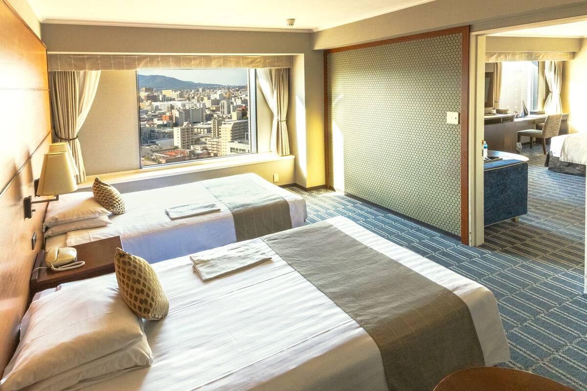 Quarto de hotel com duas camas, vista para a cidade, banheiro com espelho e escrivaninha com cadeira.