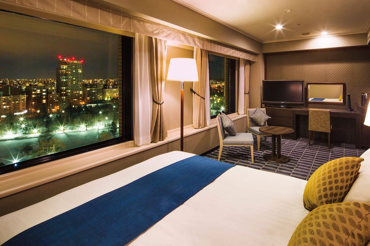 Quarto de hotel com uma cama grande, uma escrivaninha, uma cadeira, uma televisão e vista para a cidade à noite.