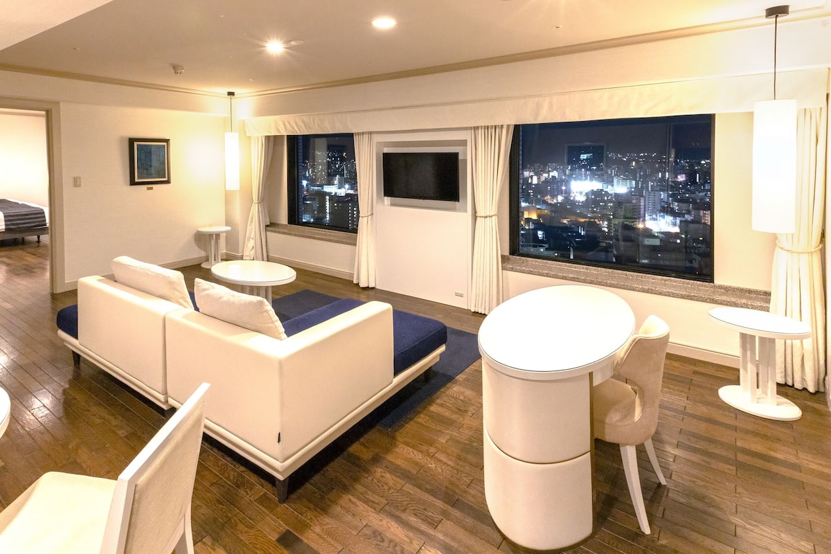 Quarto de hotel moderno com sofá, duas mesas redondas, uma TV e vista para a cidade.