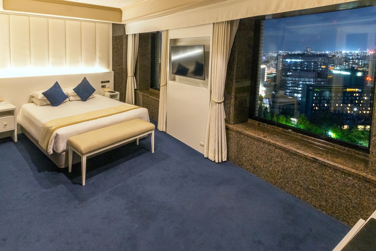 Quarto de hotel com cama, mesas de cabeceira, um banco, televisão e vista para a cidade.