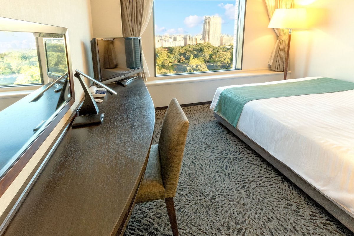 Quarto de hotel com uma cama, uma escrivaninha com computador, uma cadeira, uma janela com vista para edifícios e árvores, e um abajur.