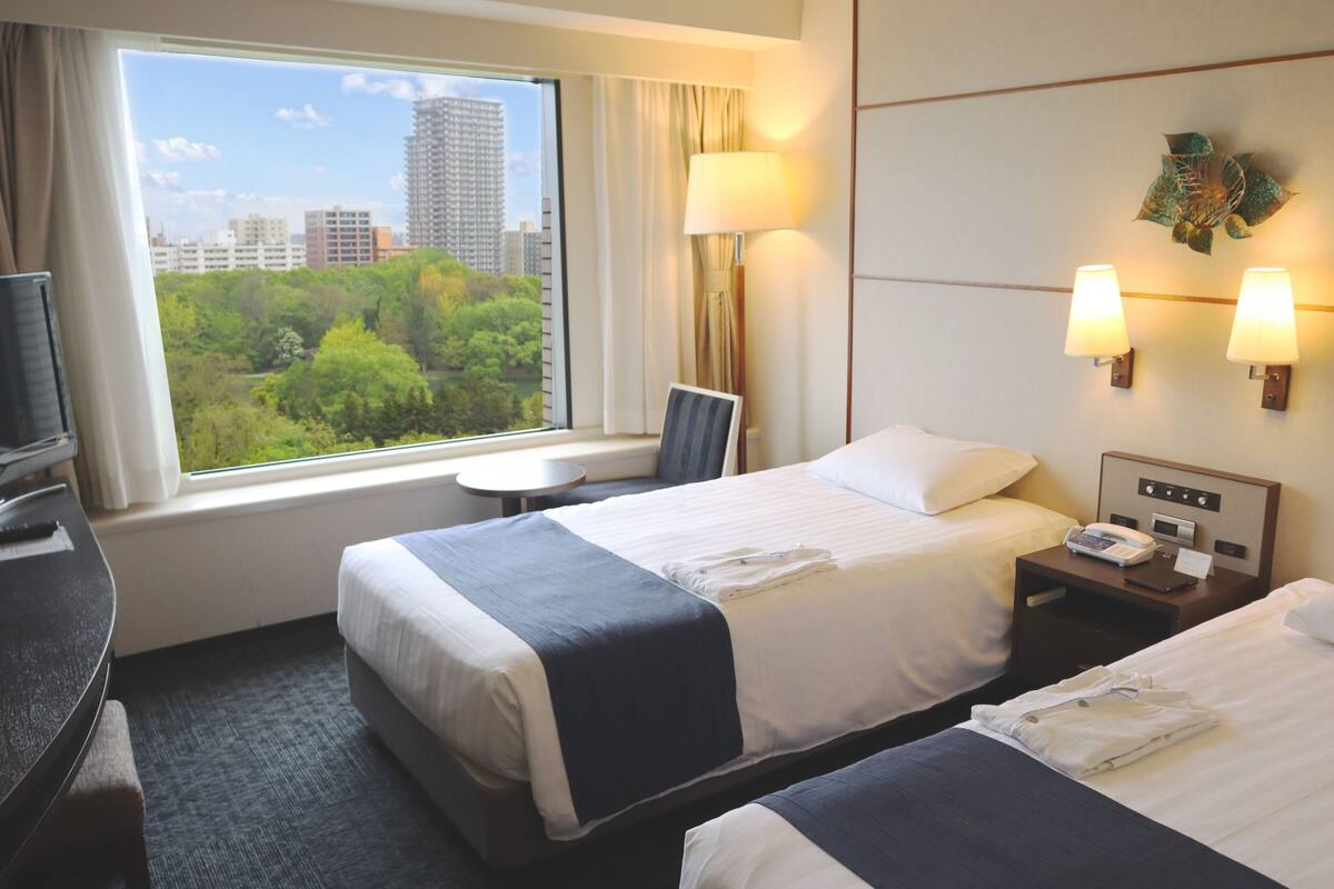 Quarto de hotel com duas camas, uma escrivaninha, uma cadeira, uma mesinha, uma janela com vista para a cidade e um telefone fixado na parede.