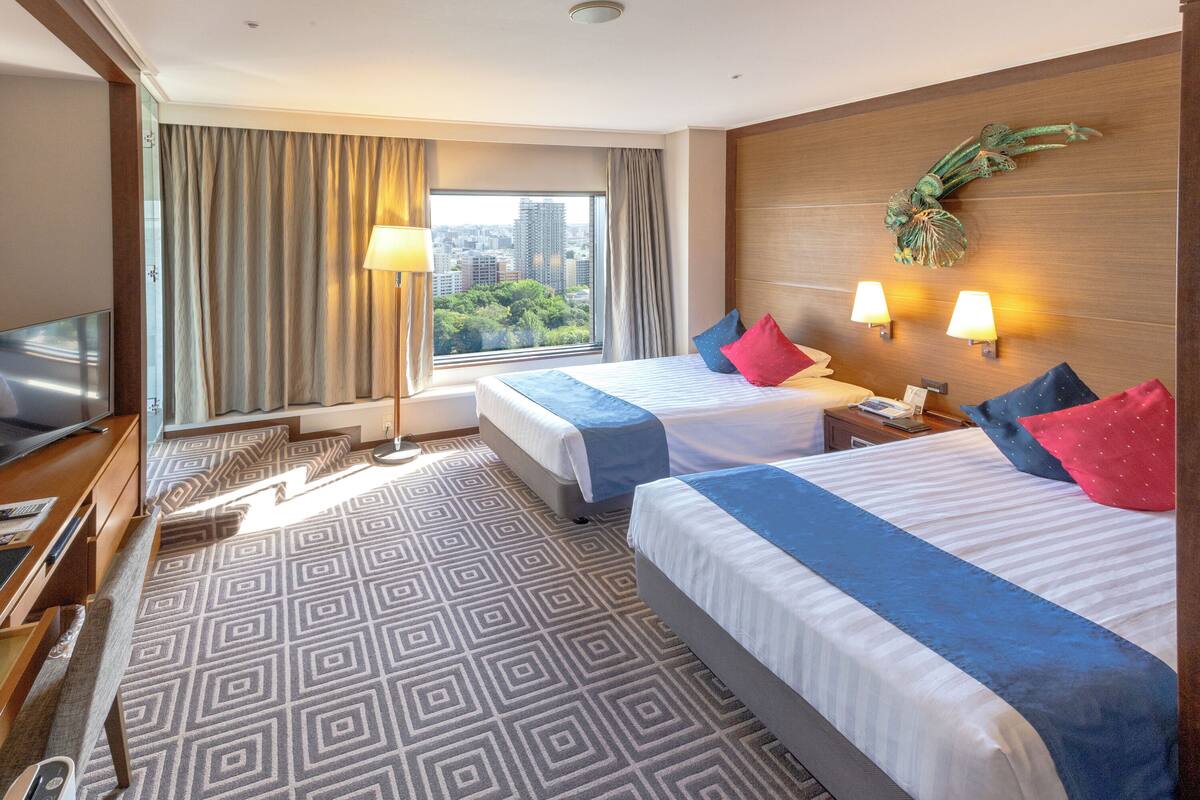 Quarto de hotel com duas camas, televisão, escrivaninha e uma janela grande com cortinas.