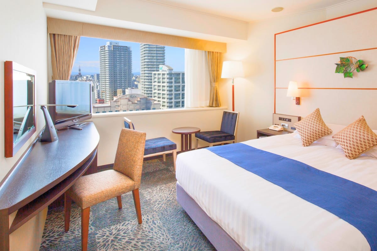 Quarto de hotel com cama, escrivaninha, cadeira e vista para edifícios da cidade.
