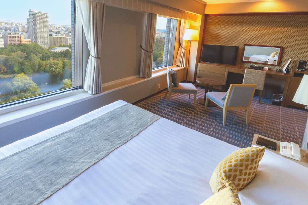 Quarto de hotel com uma cama grande, escrivaninha, cadeira, televisão e vista para a paisagem urbana e um lago.