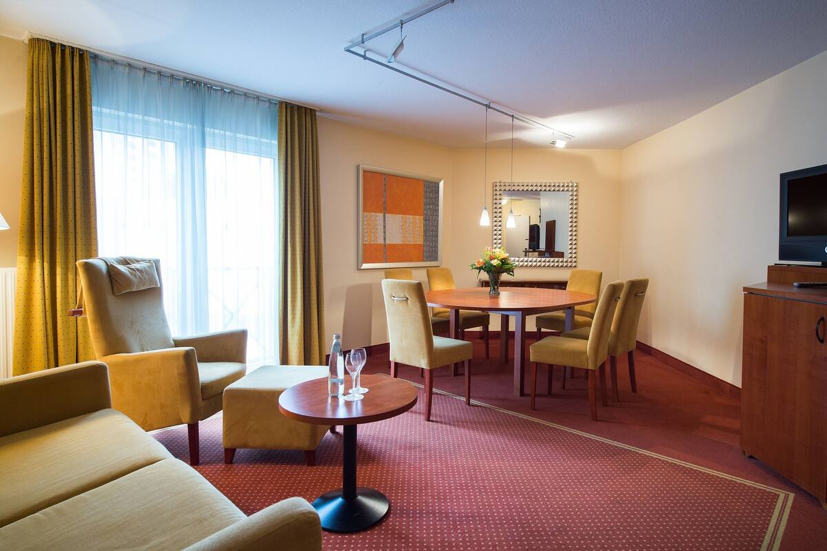Quarto de hotel com sofá, poltrona, mesa de jantar com cadeiras, televisão e espelho.