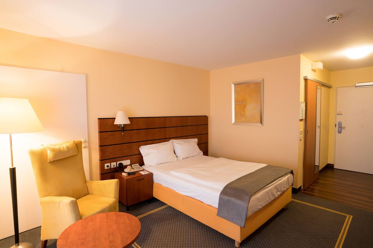 Quarto de hotel com uma cama, uma cadeira, uma mesa, um abajur, uma mesa de cabeceira e uma porta.
