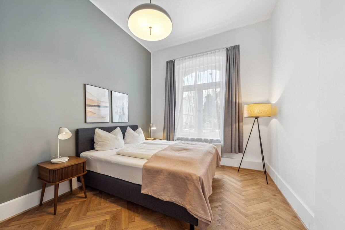 Quarto com cama, mesas de cabeceira, abajur e janela com cortinas.