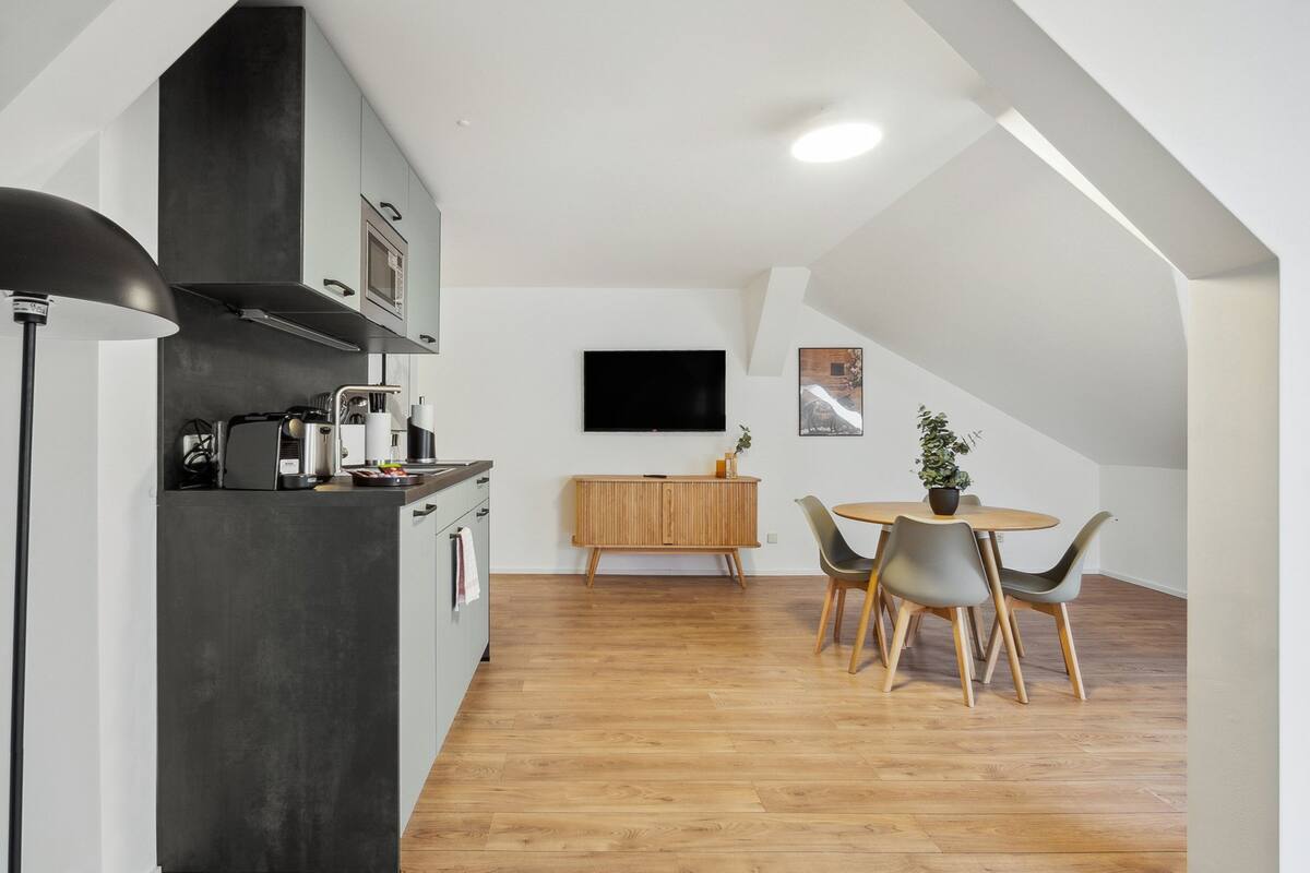 Uma cozinha moderna com forno embutido, área de jantar com mesa redonda e cadeiras, e uma TV de tela plana na parede.