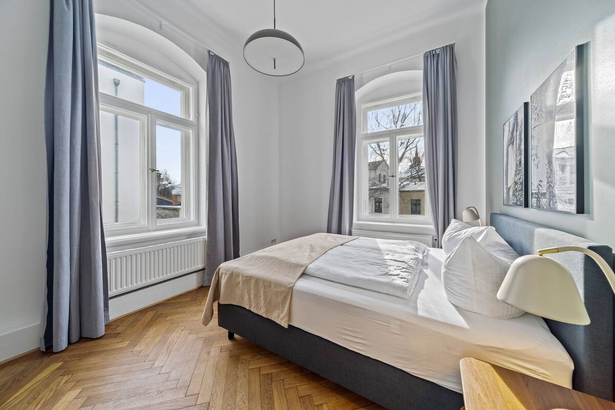 Quarto com cama, mesa de cabeceira, abajur e duas janelas com cortinas.