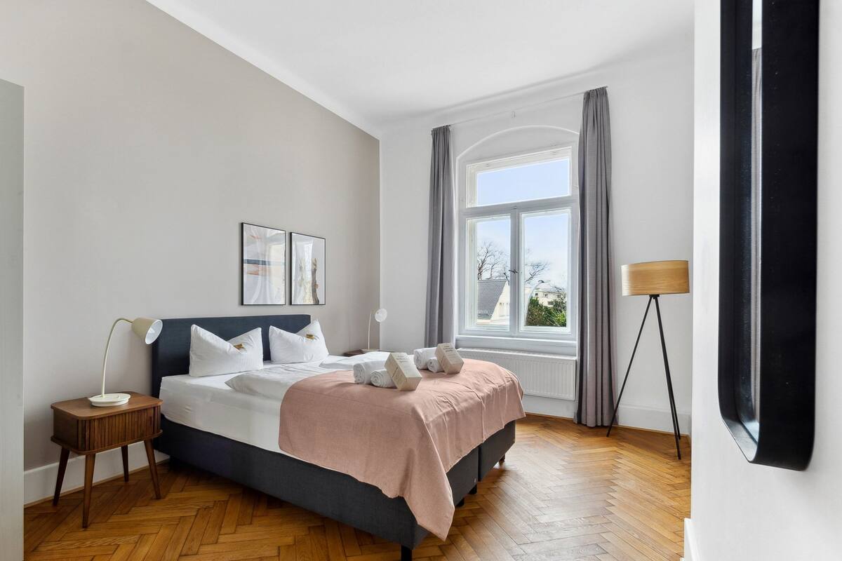 Quarto moderno com cama grande, mesa de cabeceira, abajur e janela com cortinas.