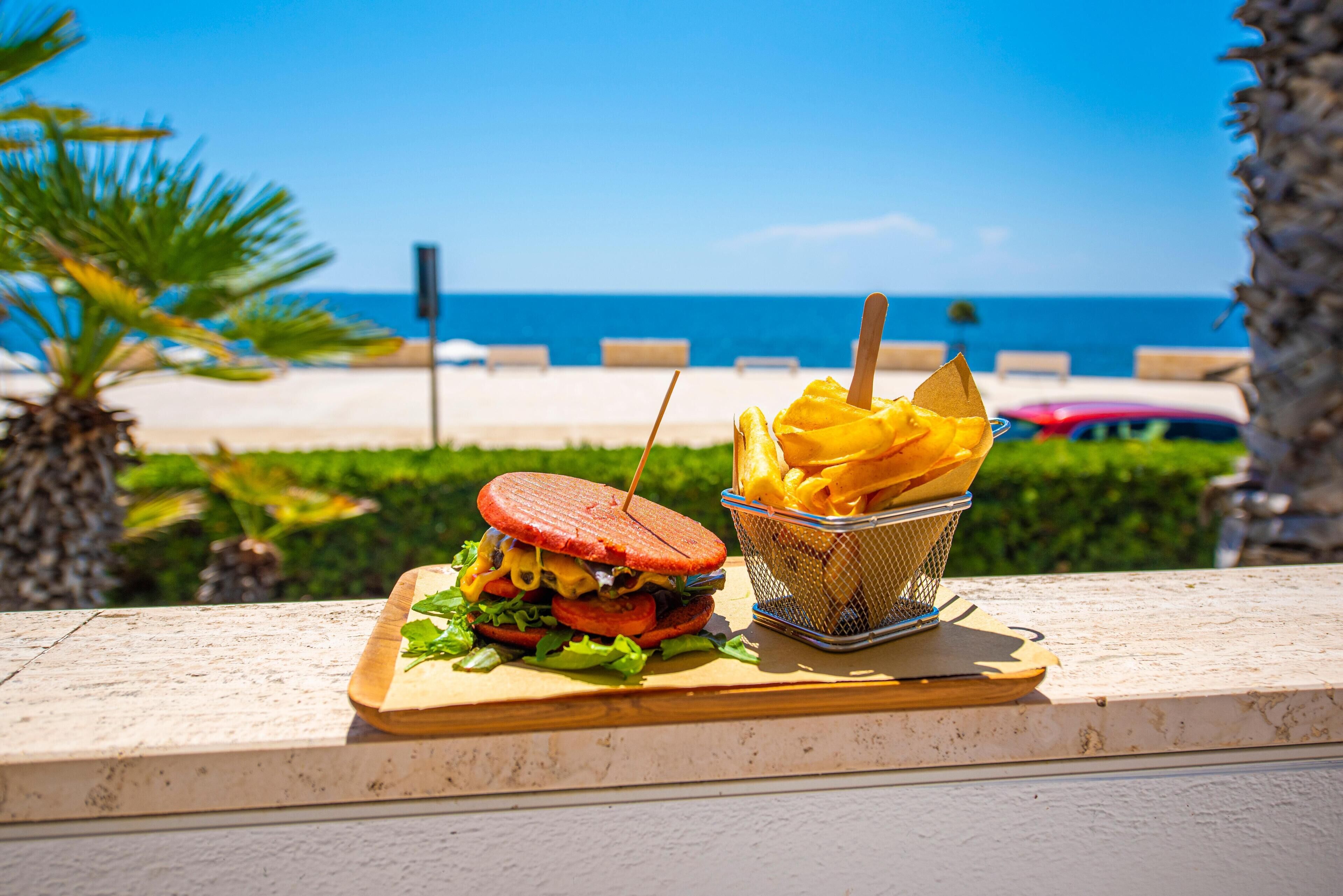 Vista da praia com um sanduíche e batatas fritas em uma tábua de madeira.