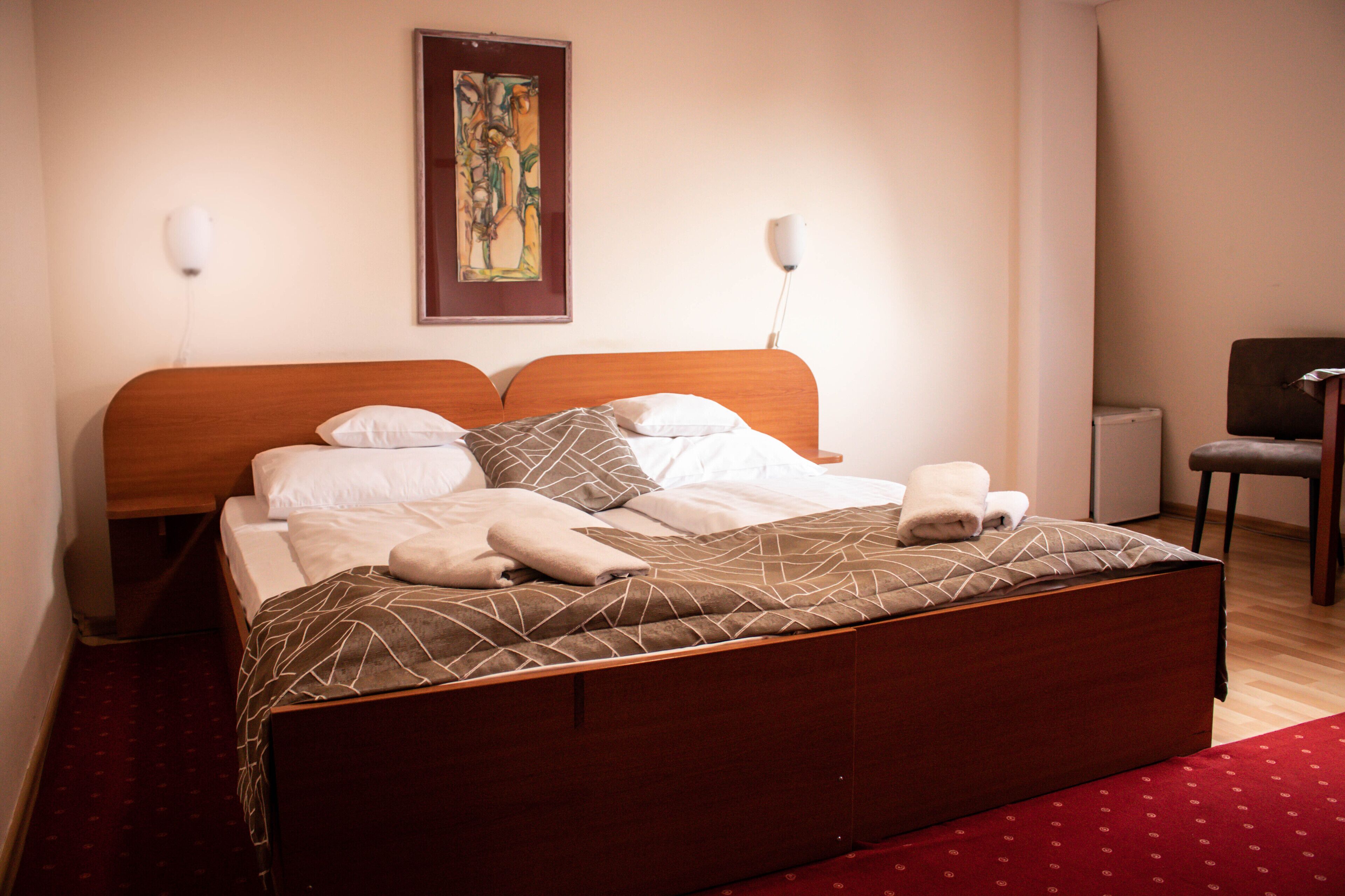 Cama bem arrumada com cabeceira de madeira, almofadas brancas com padrão e colcha vermelha. Há uma obra de arte emoldurada na parede acima da cama e uma cadeira no canto.
