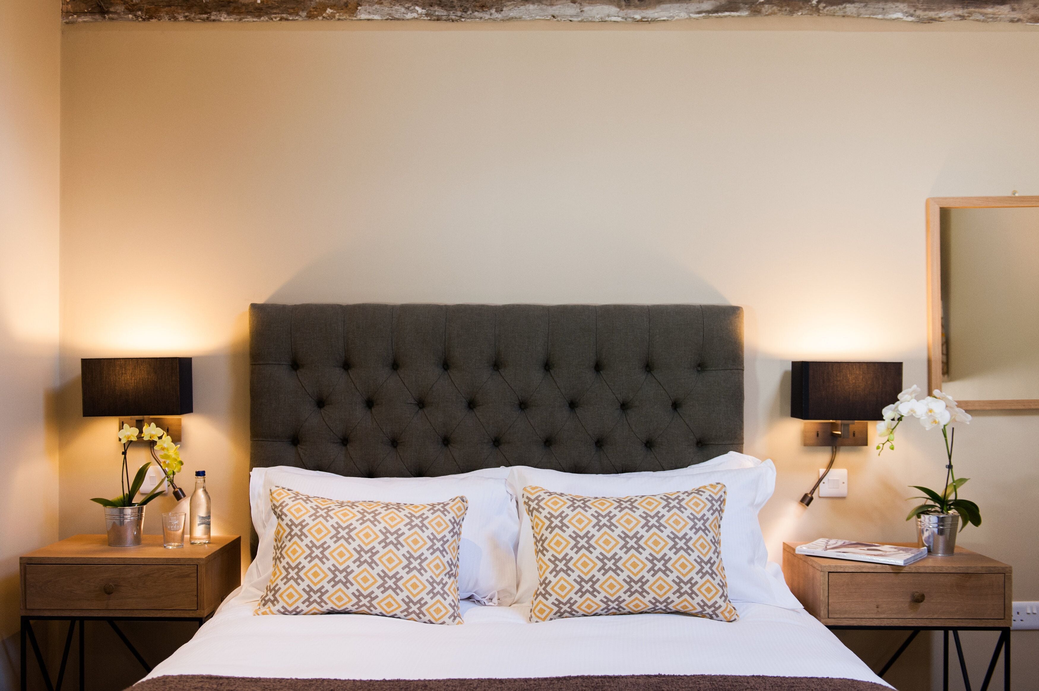 Uma cama bem arrumada com almofadas estampadas, cabeceira estofada, mesas de cabeceira com abajures e um vaso com flores.