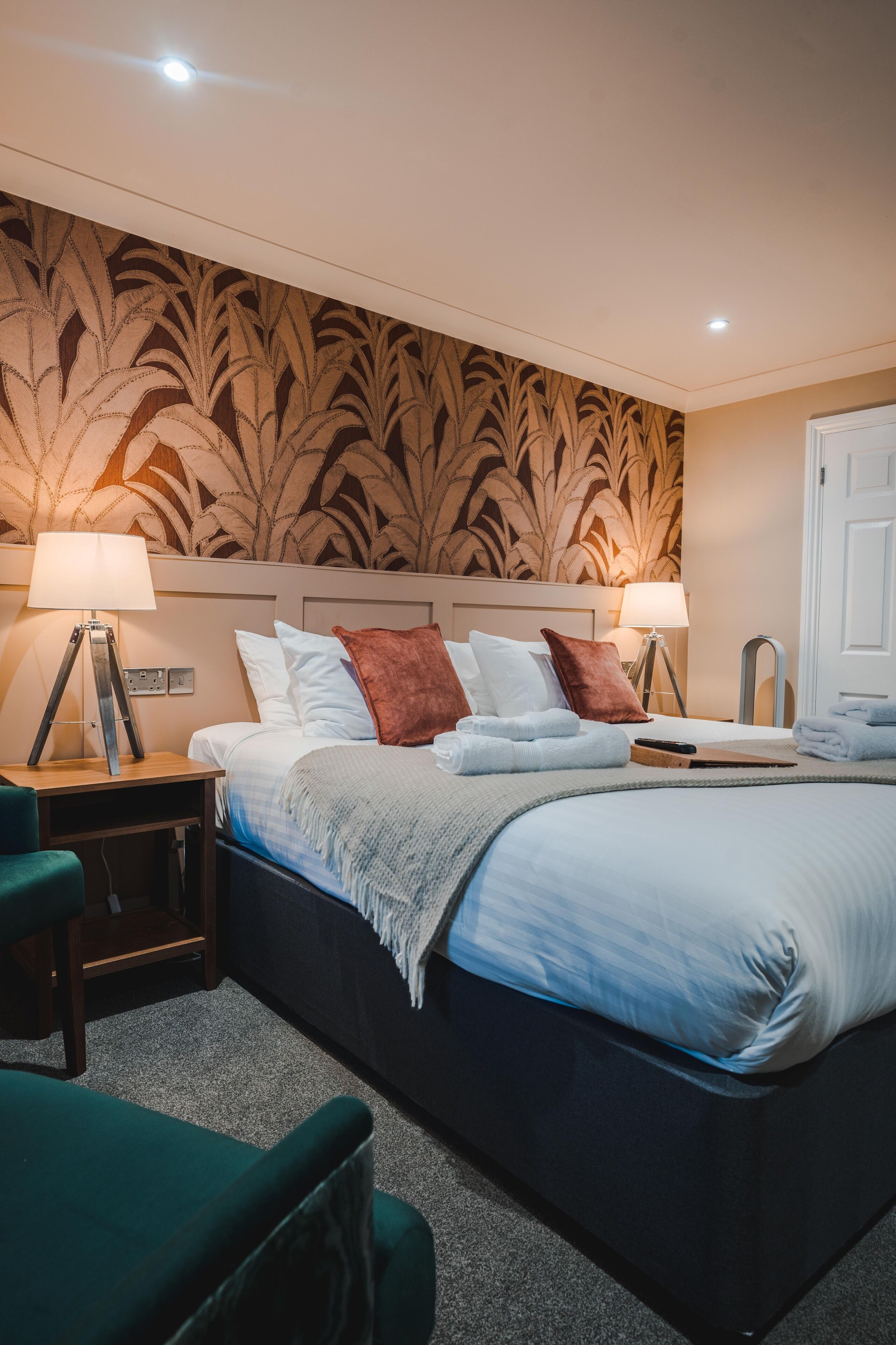 Quarto de hotel com uma cama grande, duas mesas de cabeceira, uma poltrona verde e um papel de parede estampado na parede.