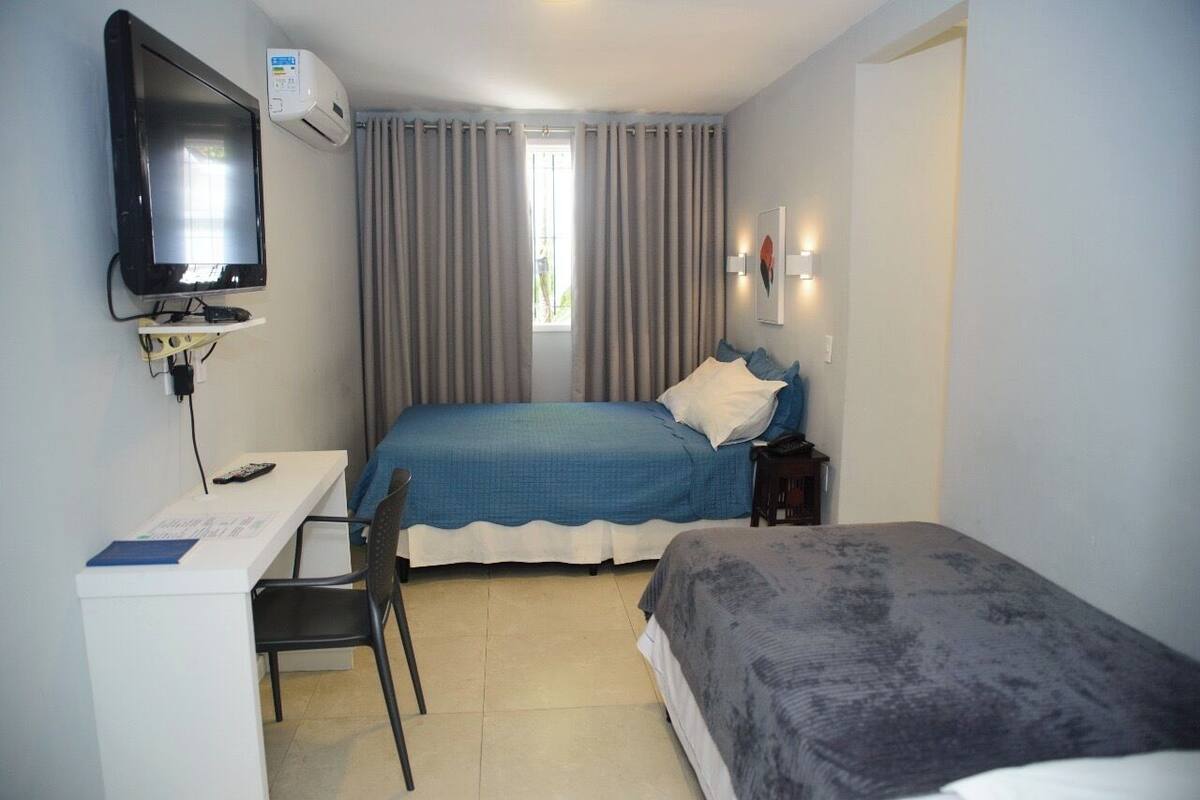 Quarto com cama, escrivaninha com cadeira, televisão e ar condicionado.