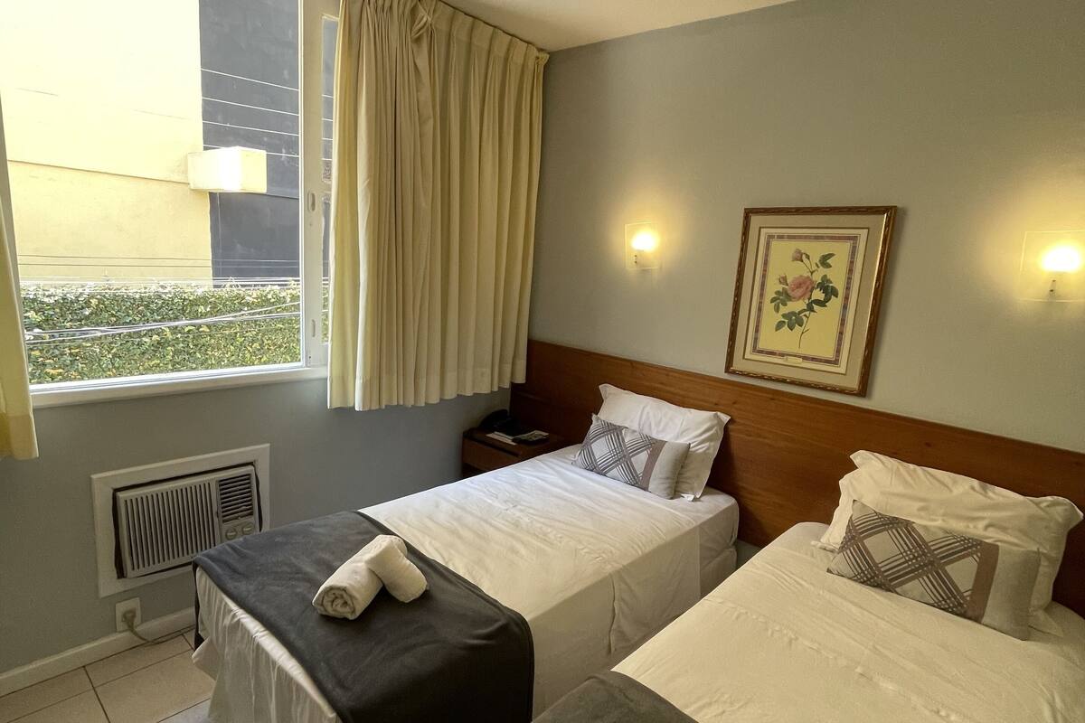 Quarto de hotel com duas camas, janela com cortinas, radiador e luminária fixada na parede.