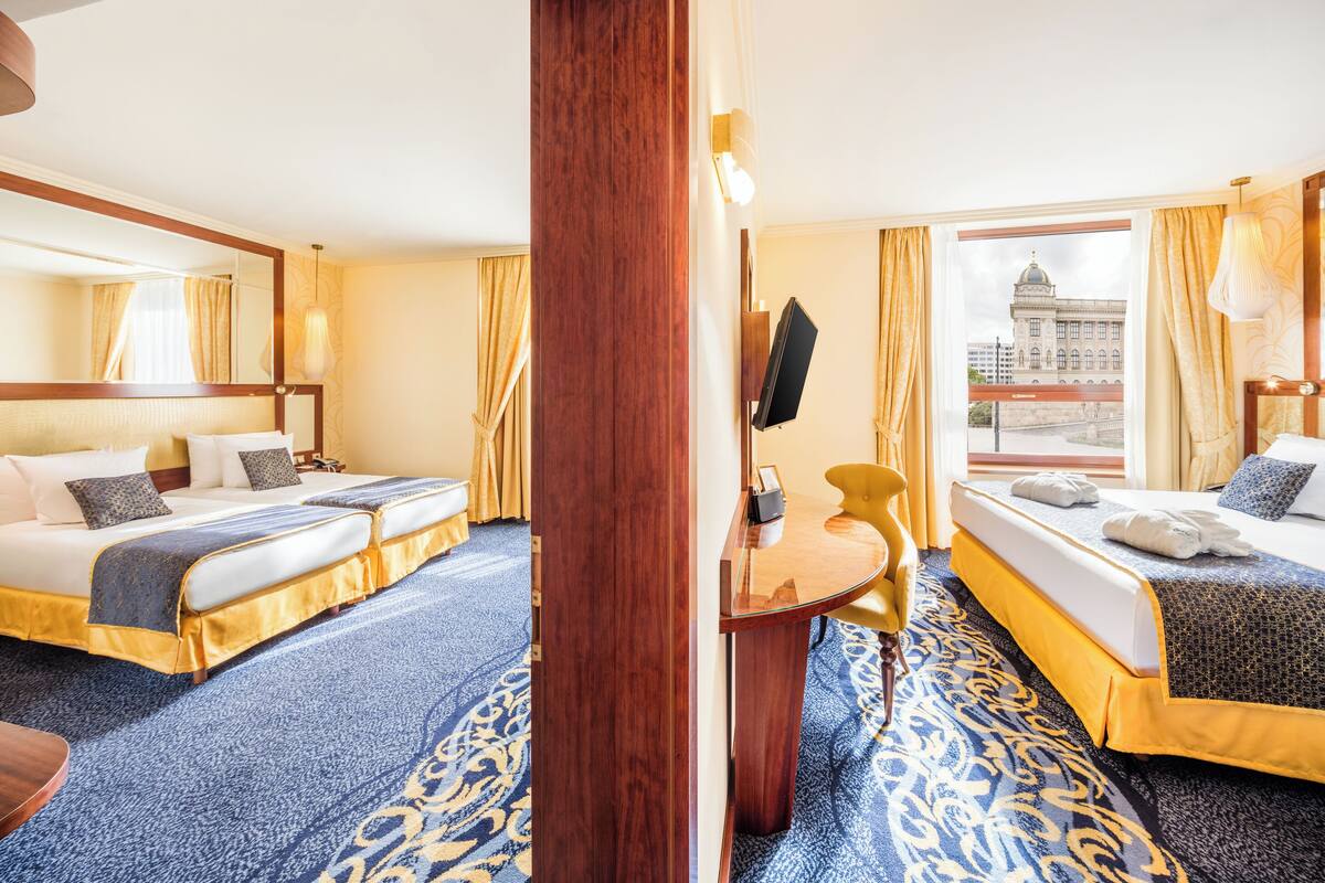 Quarto de hotel com duas camas, um pilar de madeira, uma escrivaninha e uma janela grande com vista para um edifício.